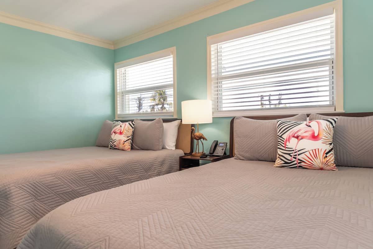 Quarto com duas camas, uma mesinha de cabeceira, uma luminária e janela com persianas.