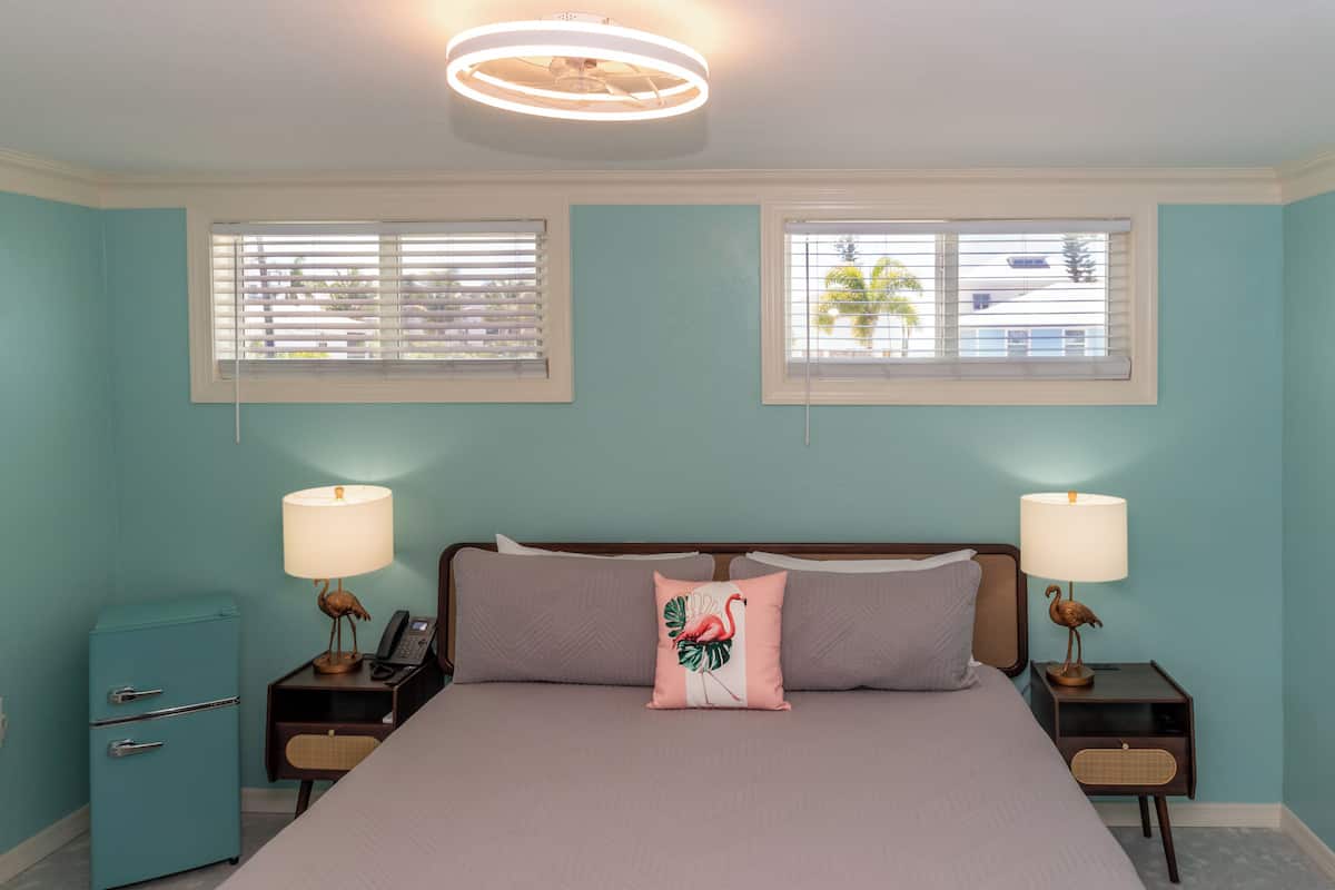 Quarto com cama, duas mesas de cabeceira com abajures, um frigorífico pequeno azul e duas janelas com persianas.