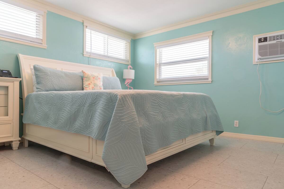 Quarto com cama, mesa de cabeceira, luminária e ar condicionado fixado na parede.