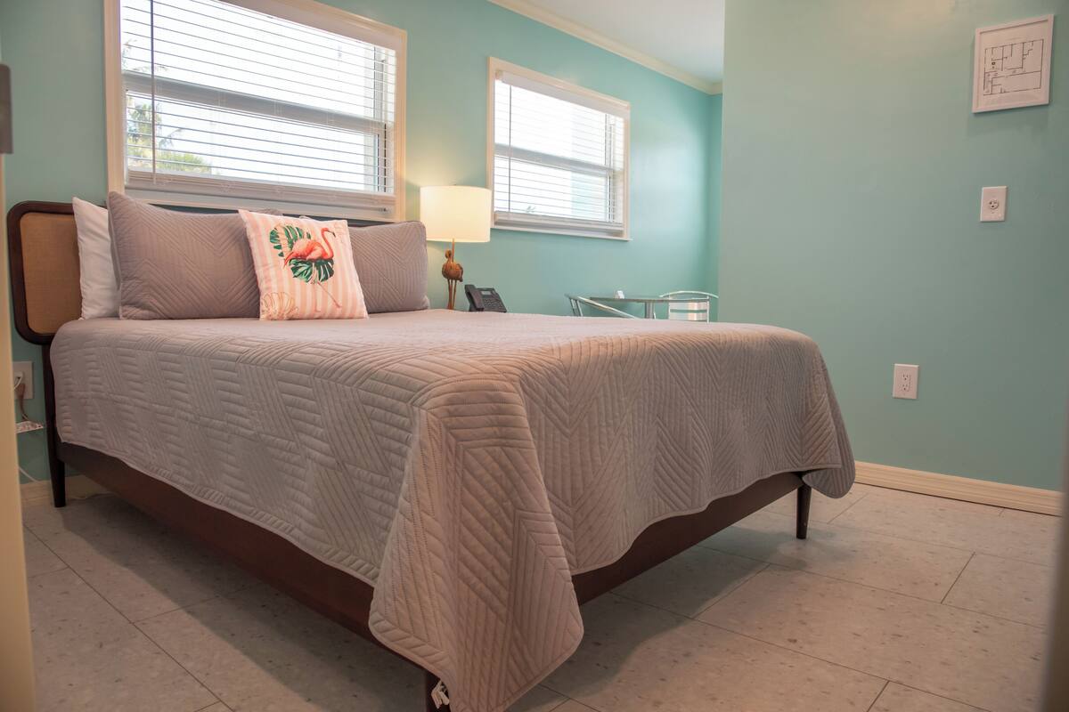 Quarto com cama, duas janelas, mesa de cabeceira e luminária.