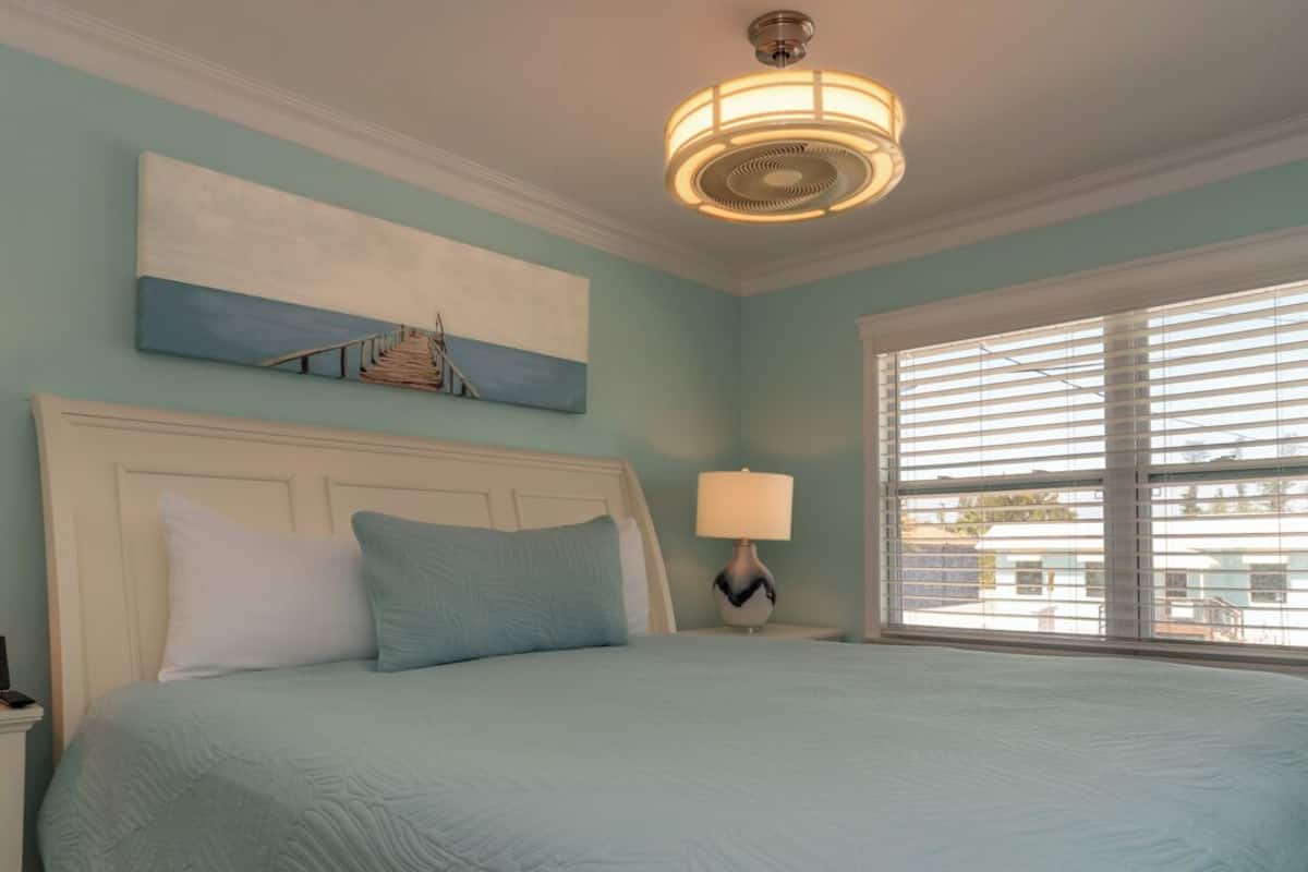 Um quarto com cama, mesa de cabeceira, luminária, ventoinha no teto, janela com persianas e um quadro na parede.
