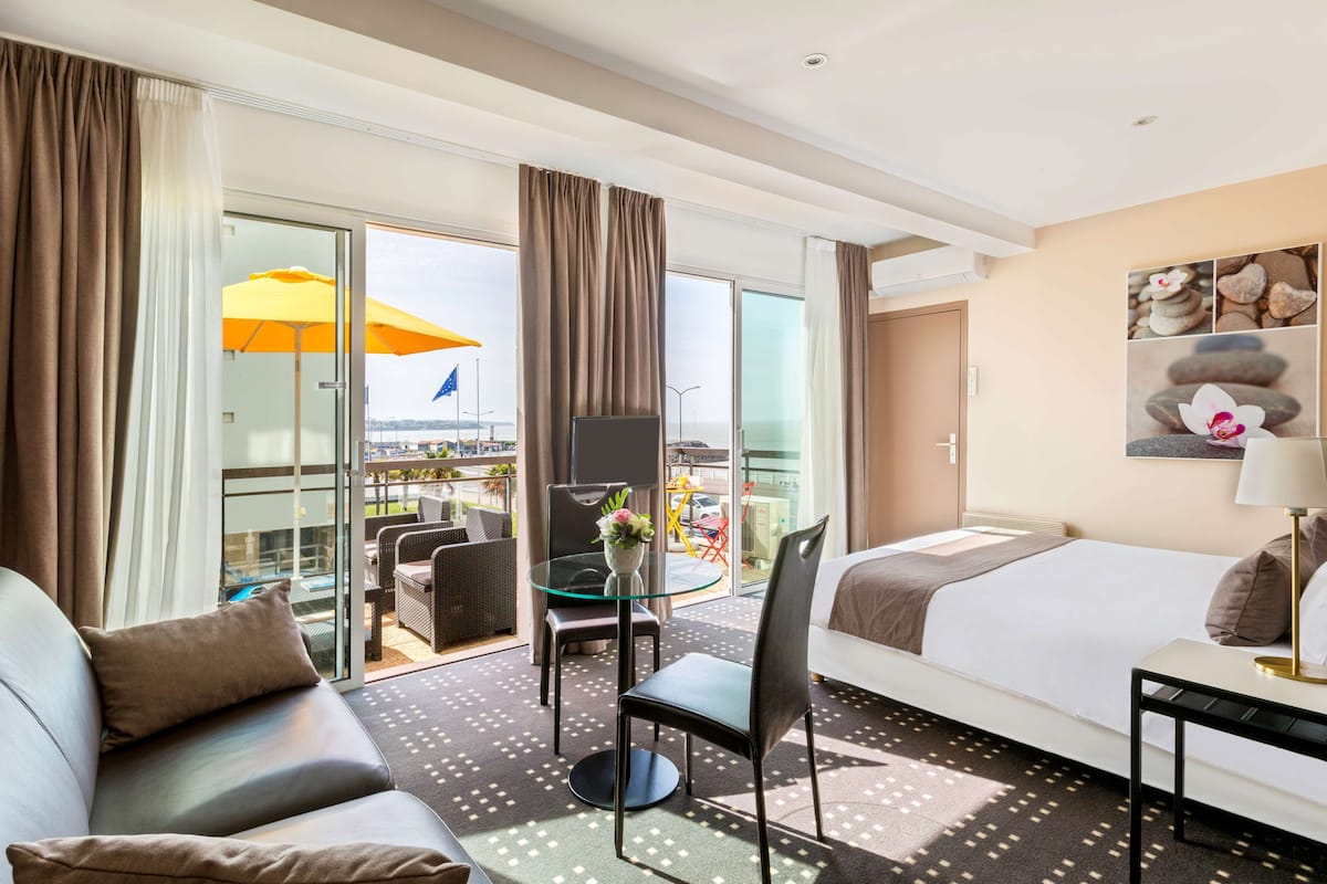Quarto de hotel com cama, sofá, mesa de jantar e varanda com guarda-sol.