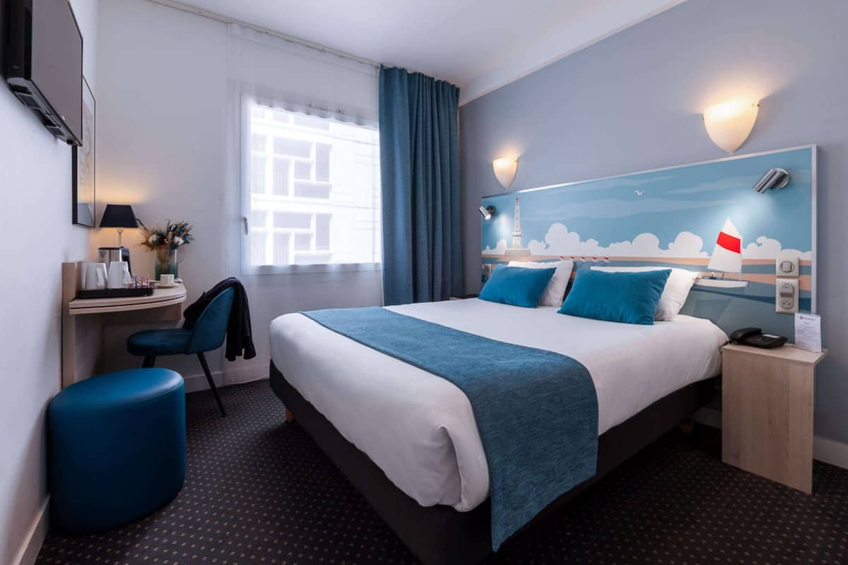 Quarto de hotel com uma cama, escrivaninha, cadeira, mesa de cabeceira e uma luminária decorativa na parede.