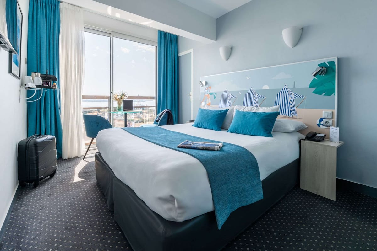 Quarto de hotel moderno com uma cama grande, vista da cidade, uma secretária pequena e uma cadeira.