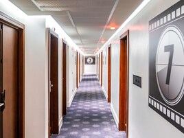 Corredor de hotel com piso carpeteado, portas de madeira e um grande número 7 na parede.