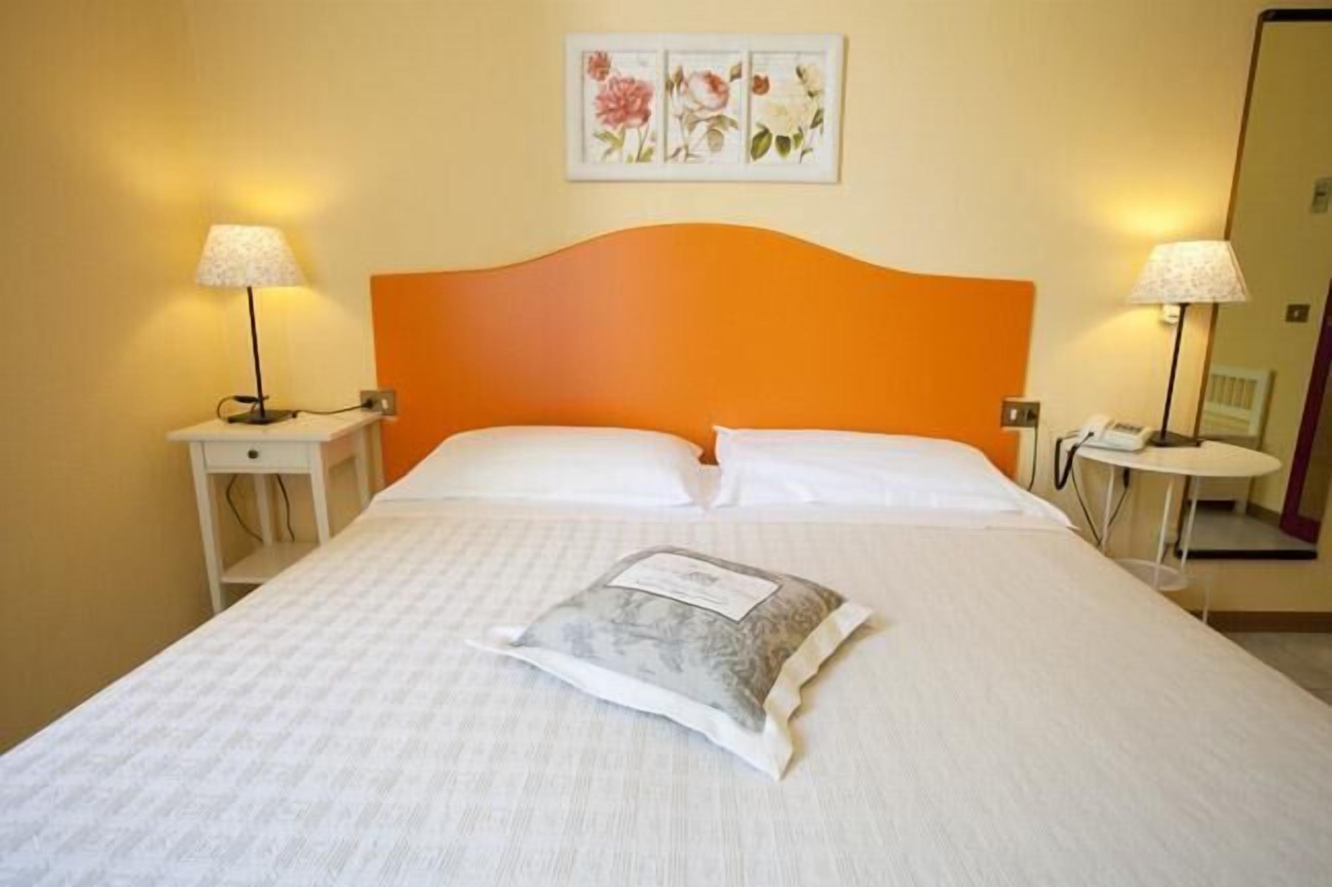 Cama bem arrumada com lençóis brancos, um travesseiro e um travesseiro decorativo. Há duas mesas de cabeceira com abajures e um quadro acima da cama.