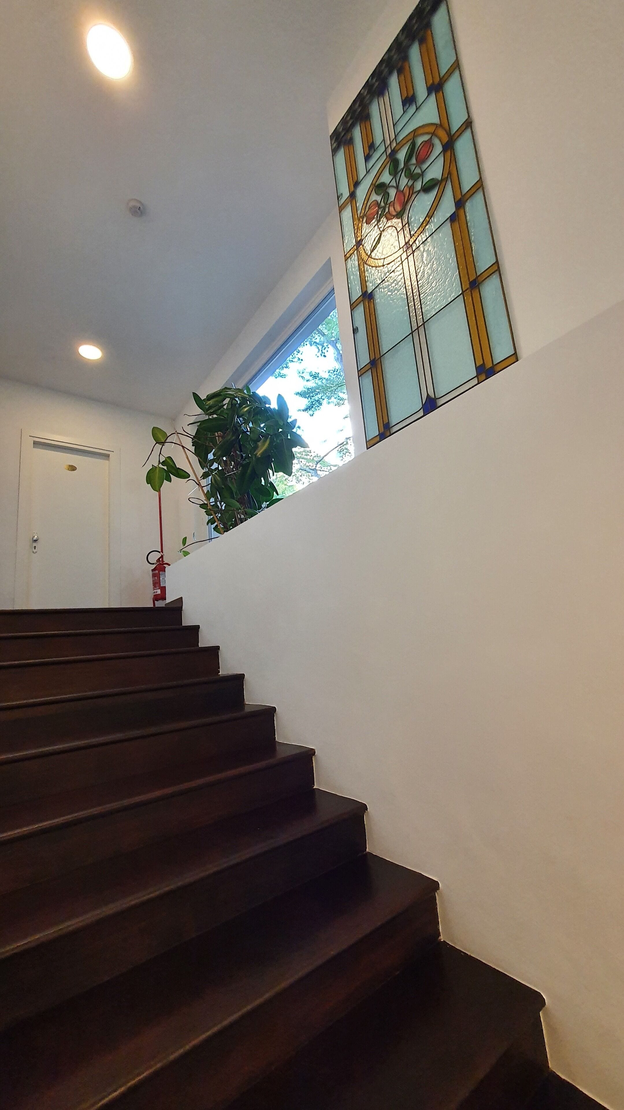 Uma escadaria com degraus de madeira leva a um patamar, onde se encontra um extintor de incêndio e uma janela com vitrais.