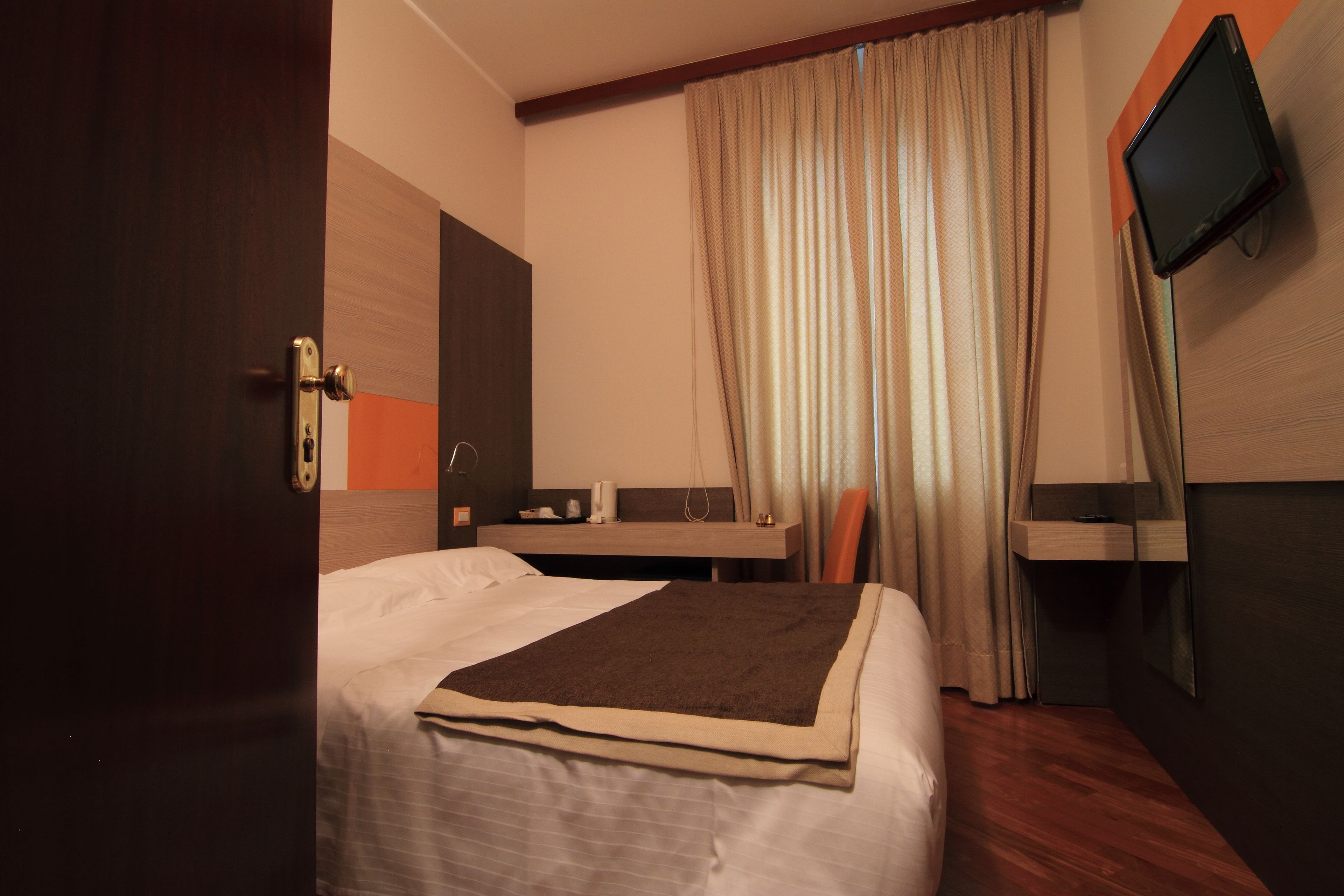 Quarto de hotel com cama, escrivaninha, cadeira, televisão e janela com cortinas.