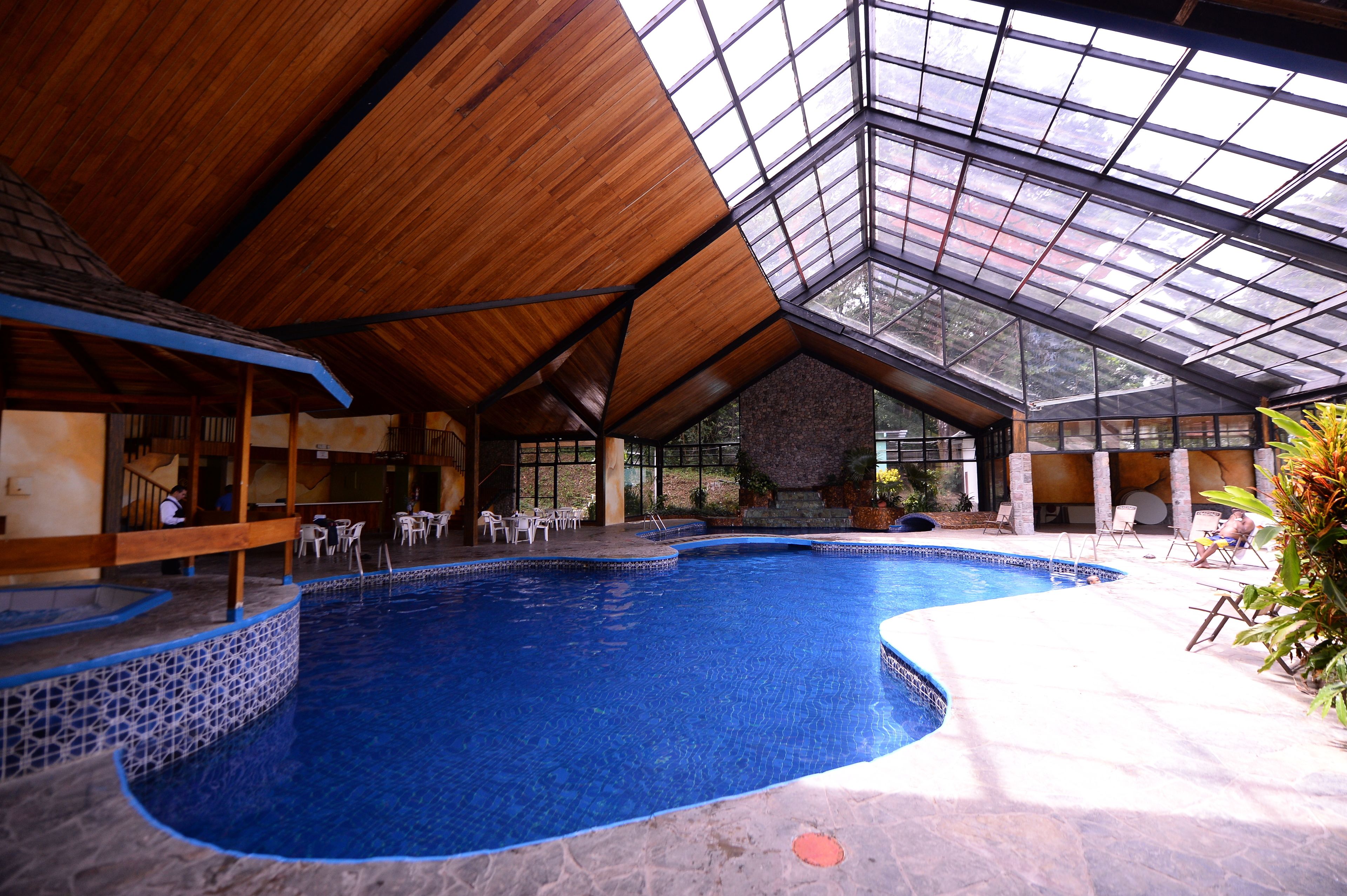 Piscina interna com teto de madeira e cobertura de vidro, rodeada por espreguiçadeiras e mesas.