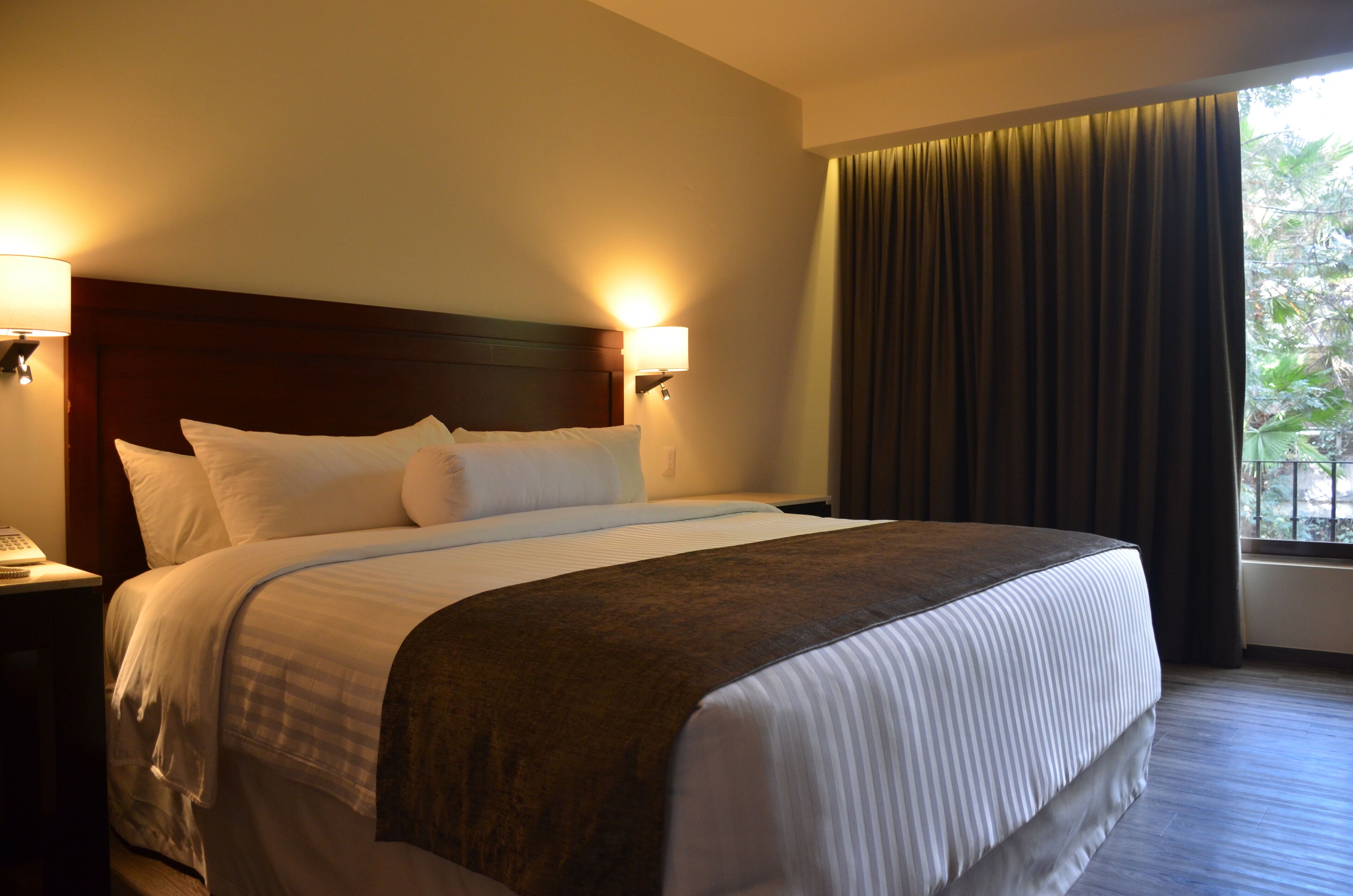 Quarto de hotel com uma cama grande, duas luminárias de cabeceira, um criado-mudo e uma janela com cortinas.