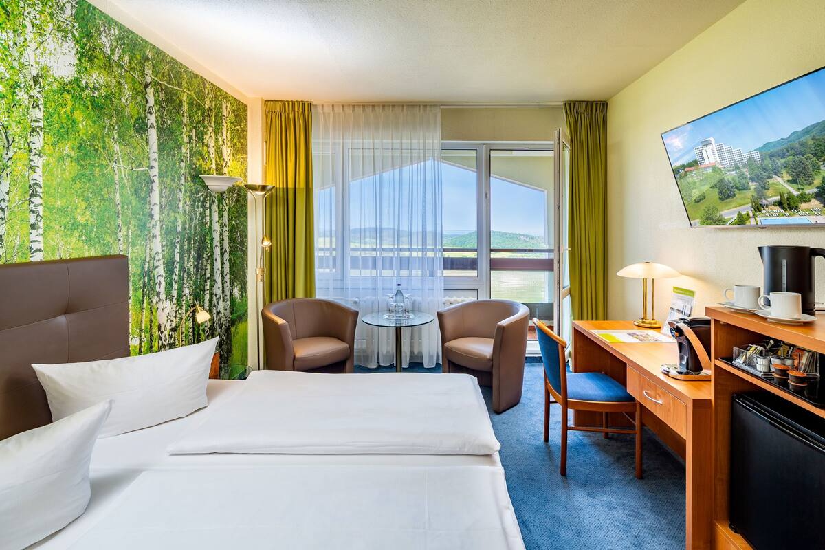 Quarto de hotel com cama, duas cadeiras, escrivaninha, televisão e vista para o exterior.