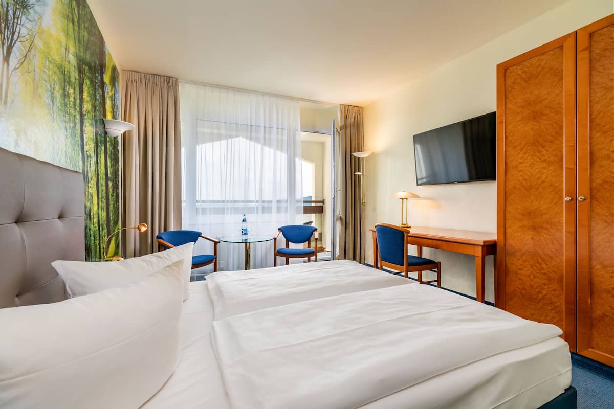 Quarto de hotel com cama, escrivaninha, duas cadeiras, televisão e uma janela grande com cortinas.