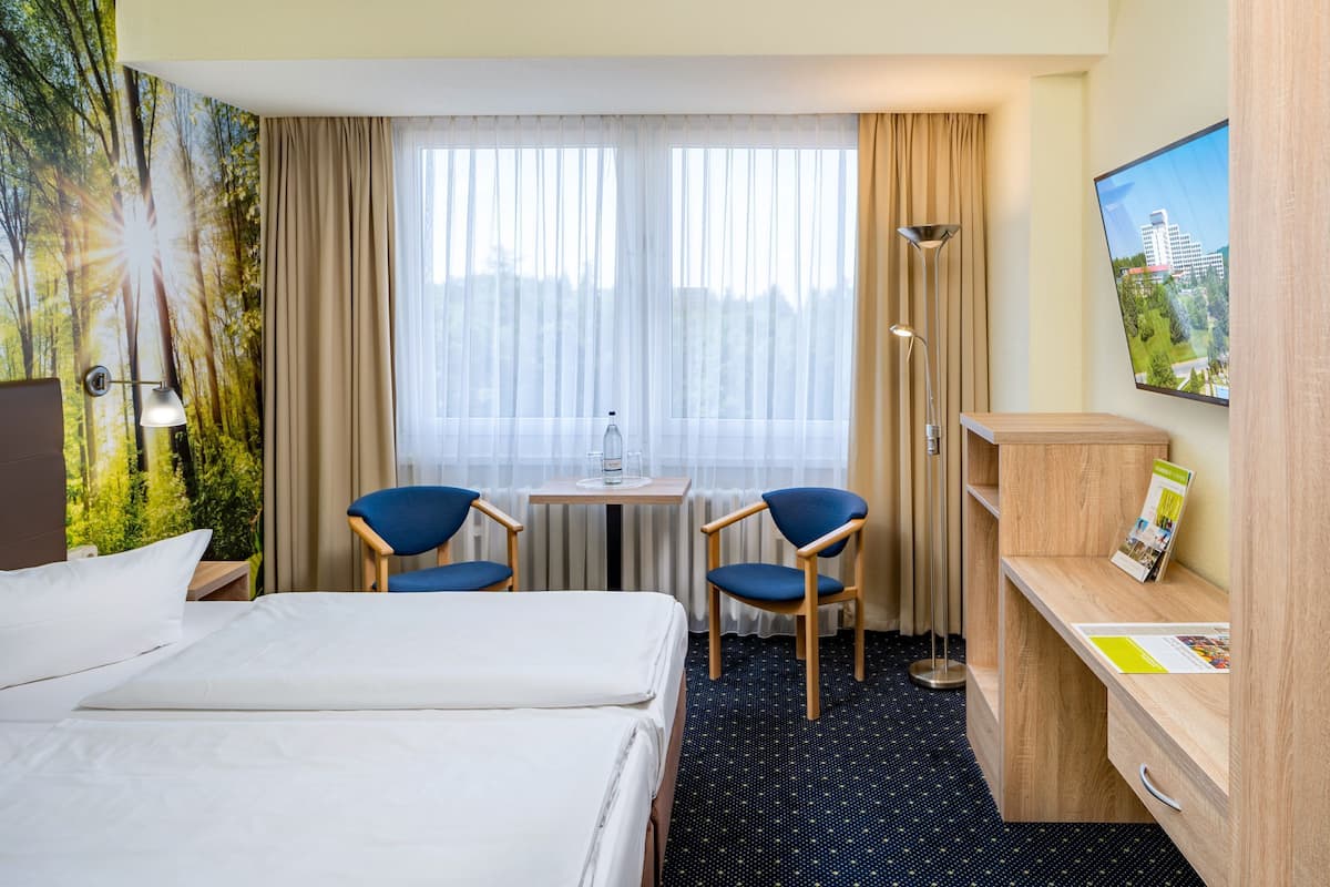 Quarto de hotel com duas camas, uma escrivaninha com cadeira, uma mesinha com garrafa e um quadro na parede.