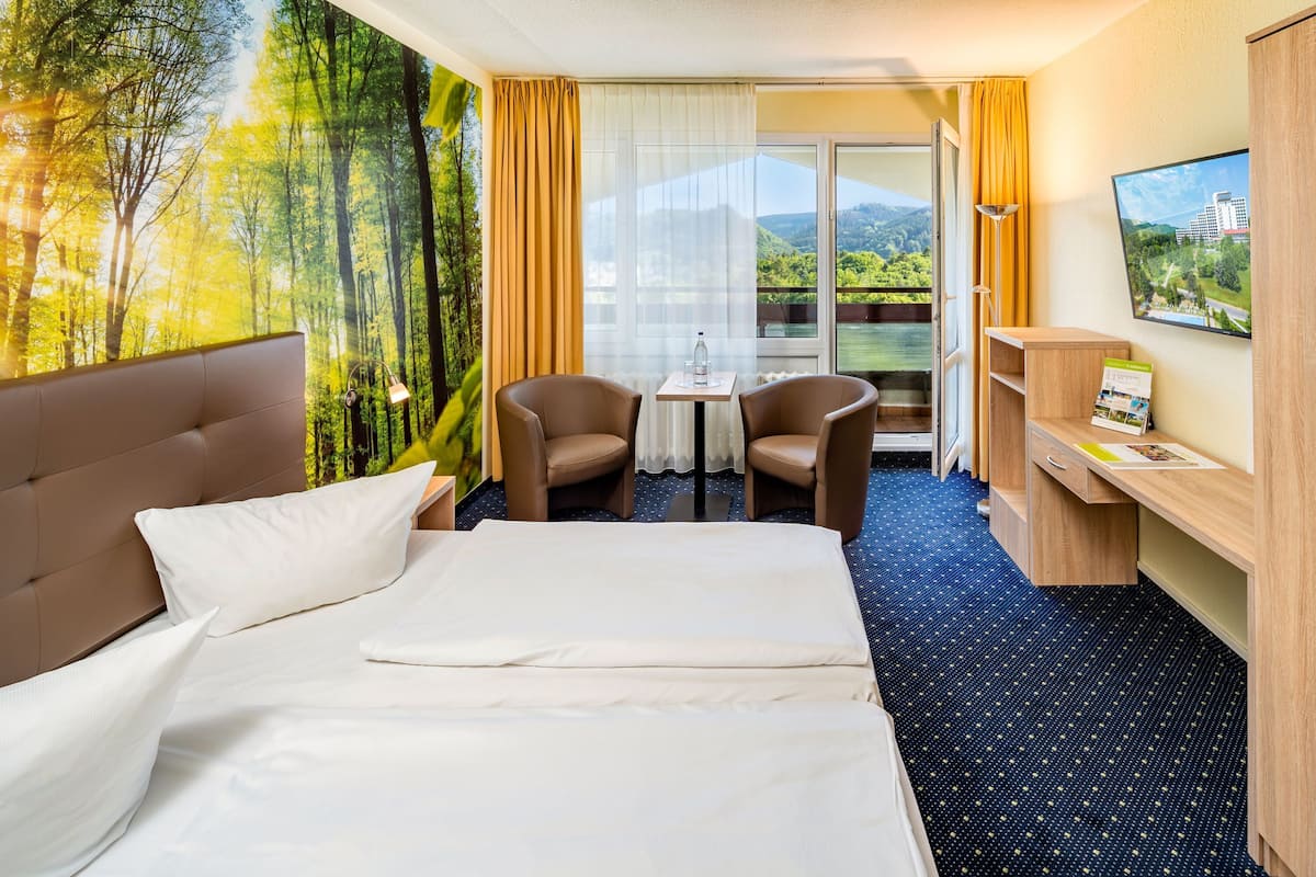 Quarto de hotel com cama, duas cadeiras, uma mesinha, escrivaninha e vista para uma floresta pela janela.