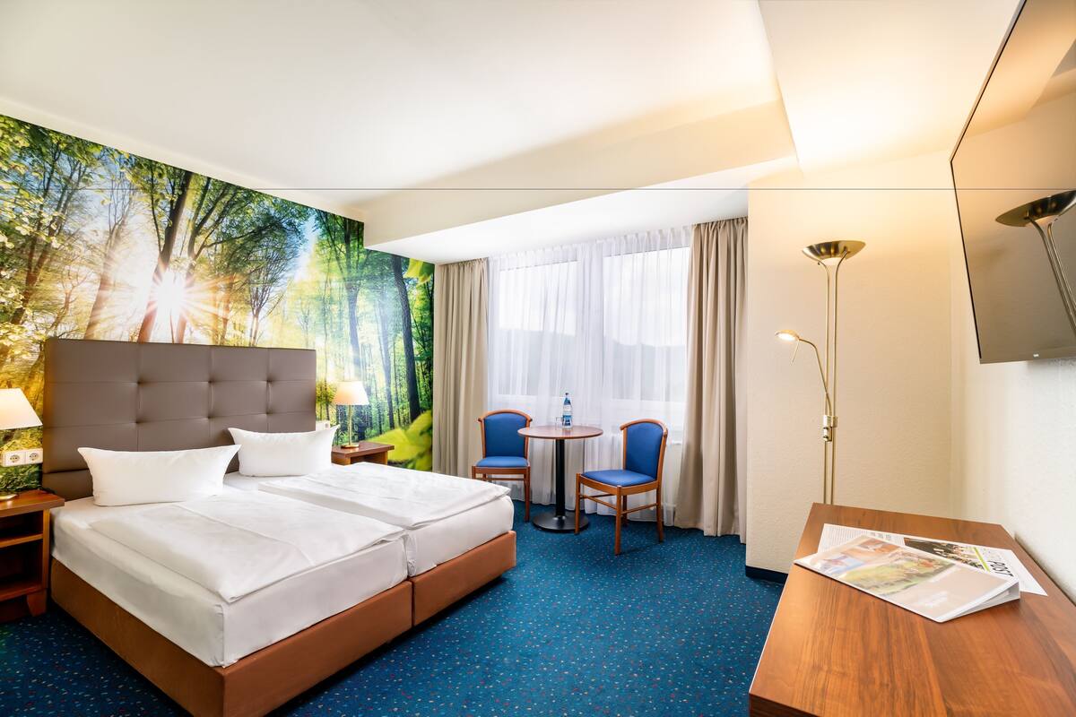 Quarto de hotel com uma cama grande, duas cadeiras, uma mesa pequena e uma janela ampla com cortinas.