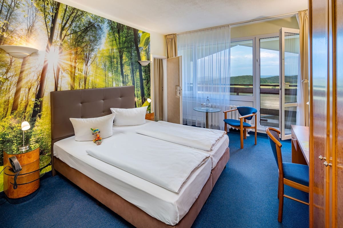 Quarto de hotel com uma cama grande, uma mesinha com cadeiras, varanda com vista e um mural de uma floresta.