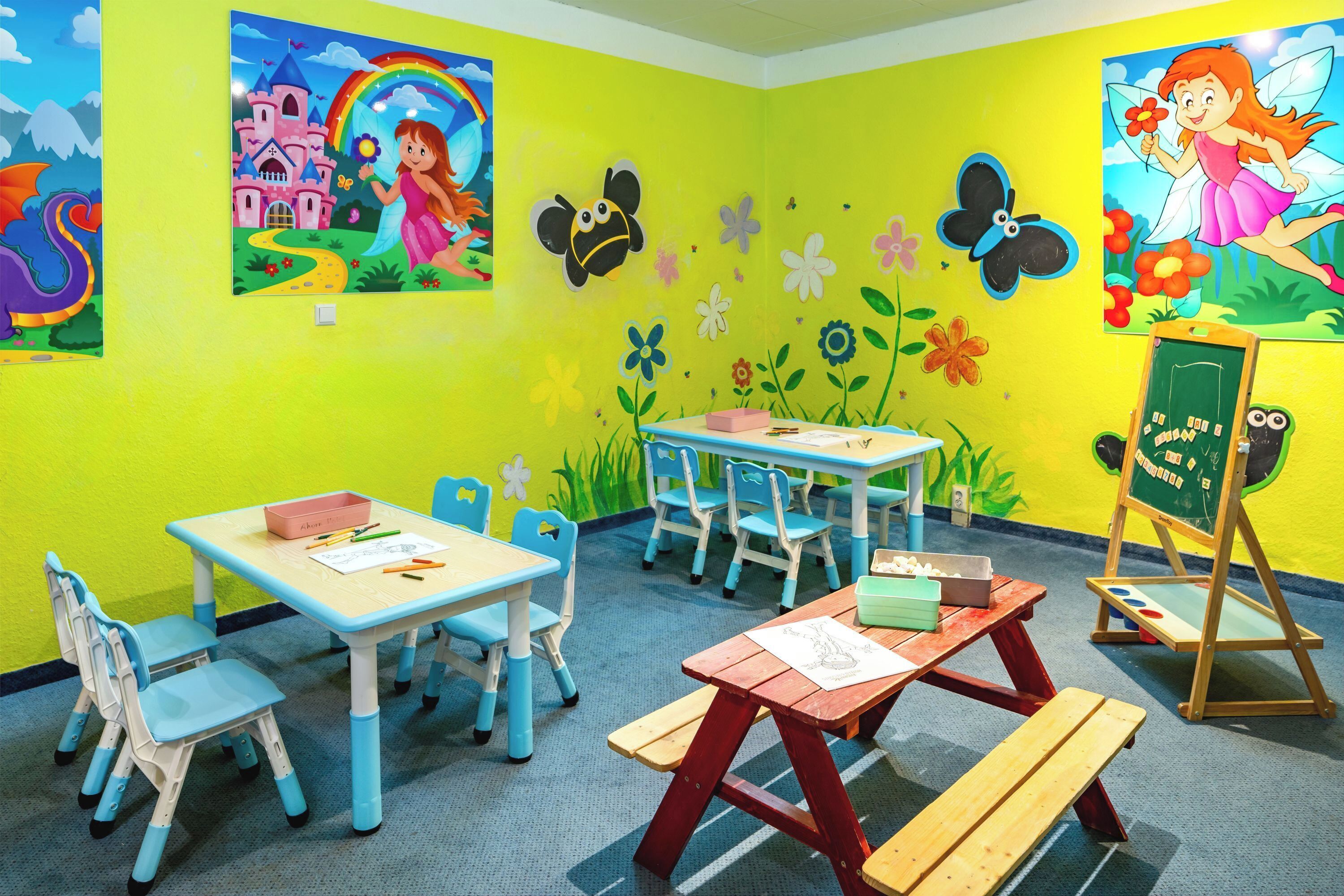 Uma sala de brinquedos infantil com paredes coloridas, um quadro negro e diversas mesas e cadeiras para brincar.