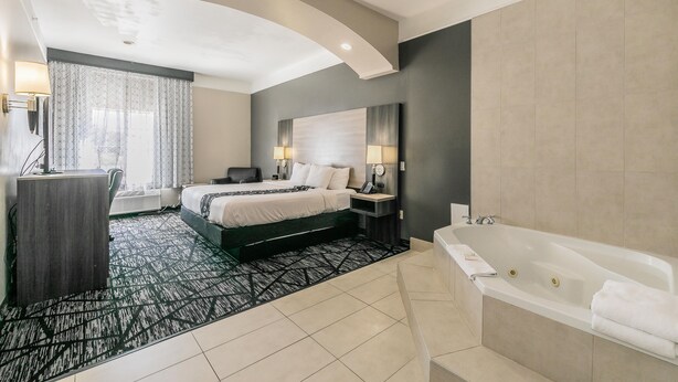 Quarto de hotel moderno com uma cama grande, uma escrivaninha, uma lâmpada de mesa, uma mesa de cabeceira, televisão fixada na parede, uma janela ampla com cortinas e uma banheira.