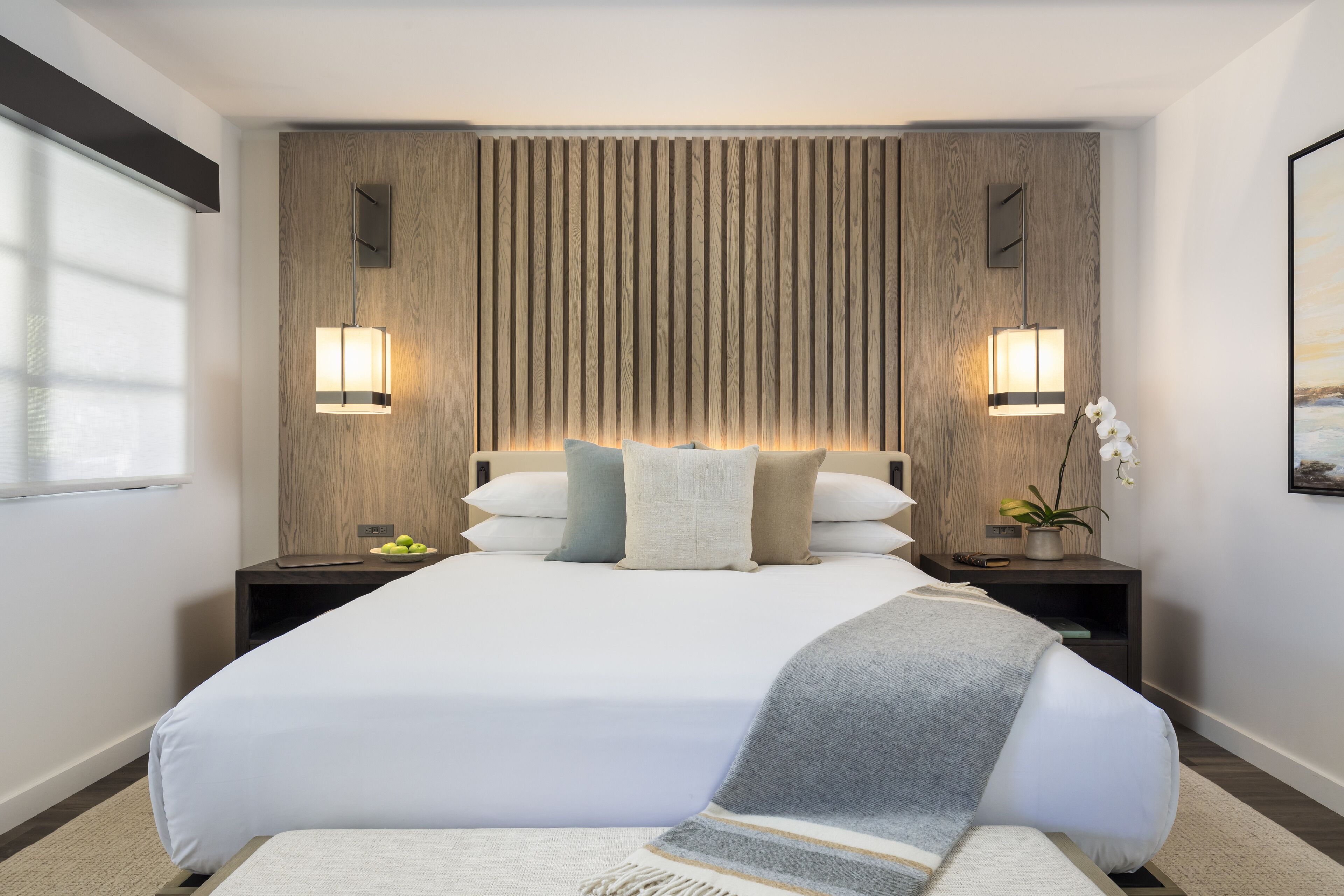 Quarto de hotel moderno com uma cama grande, painel de parede de madeira, duas mesas de cabeceira com abajures e uma pequena planta em vaso.