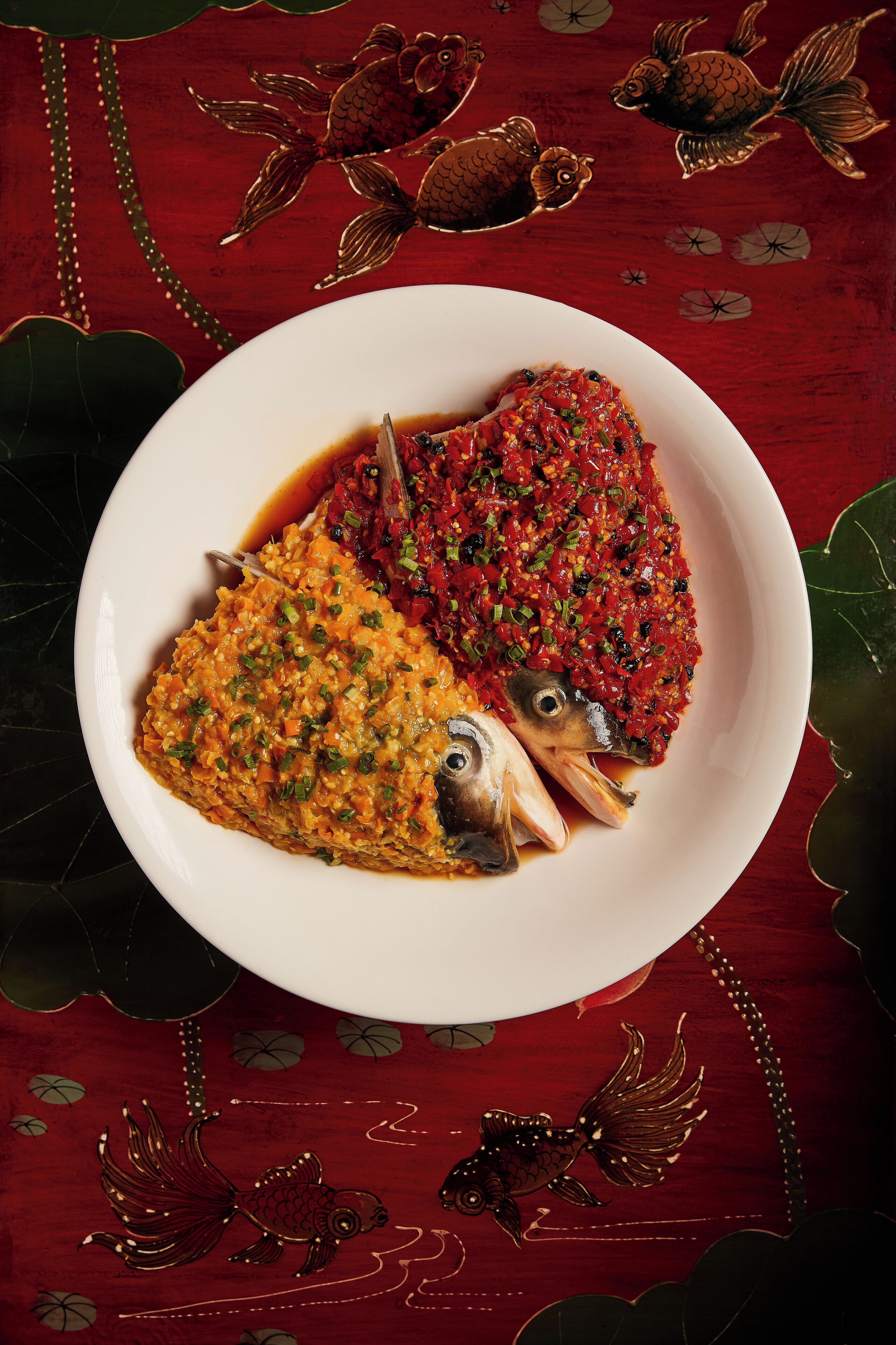 Prato branco com dois peixes inteiros e molho vermelho sobre toalha de mesa vermelha com motivos de peixes.