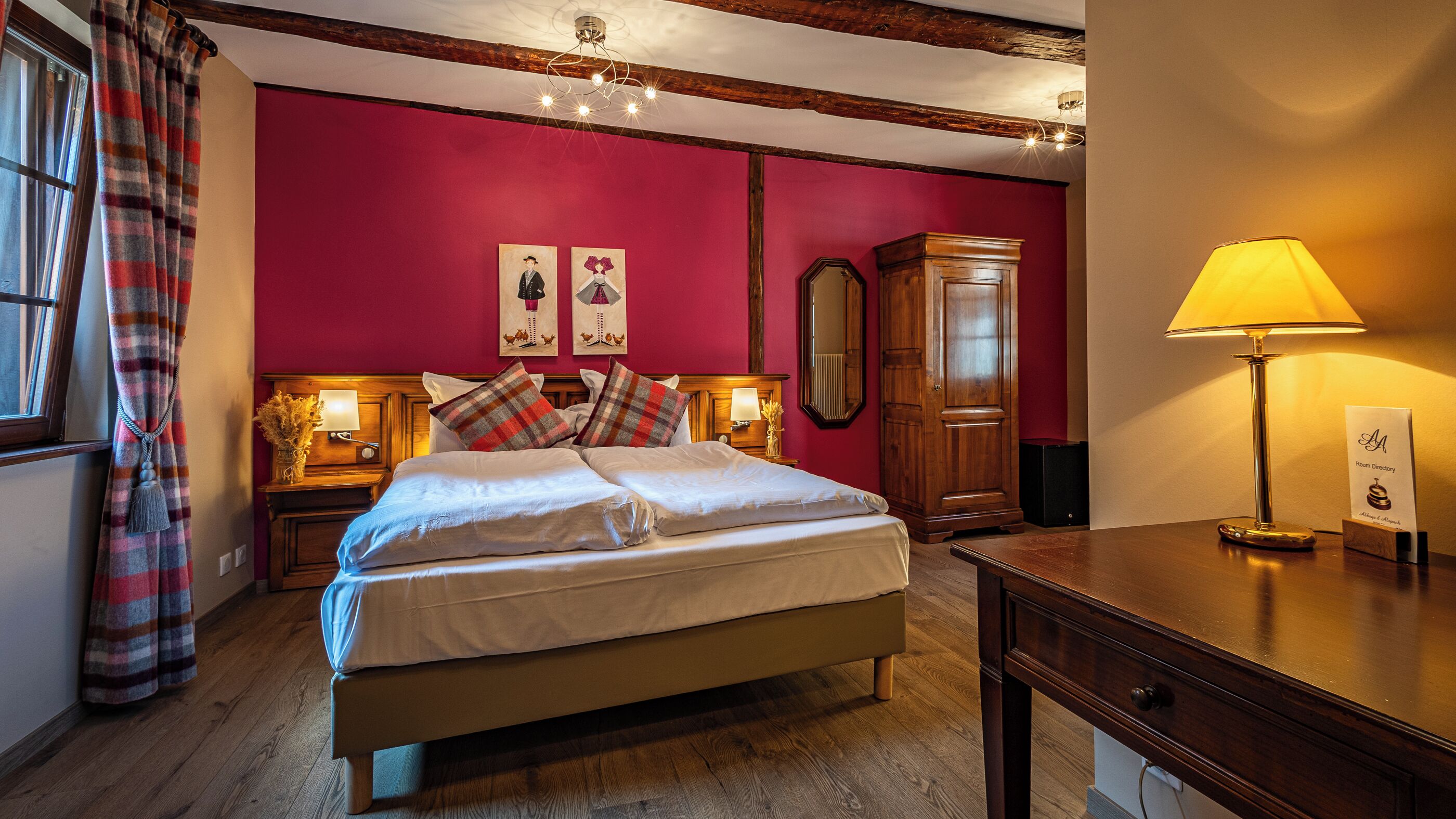 Quarto de hotel com cama, luminárias de cabeceira, mesa de cabeceira de madeira, espelho, guarda-roupa de madeira e placa indicando o número do quarto.