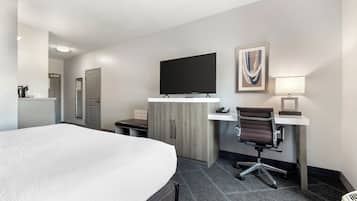 Quarto de hotel com uma cama grande, uma escrivaninha com um computador, uma cadeira, uma televisão e um quadro na parede.
