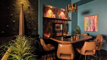 Uma área de bar com uma mesa redonda, quatro banquetas, uma parede decorativa com padrão geométrico, um pilar alto e uma luminária pendente.