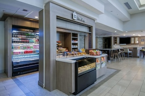 Um saguão de hotel com refrigerador de bebidas geladas, um balcão de lanches e uma área de estar com mesas e cadeiras.