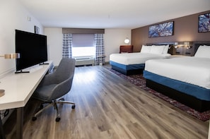 Quarto de hotel com escrivaninha, duas camas, televisão e janela com cortinas.