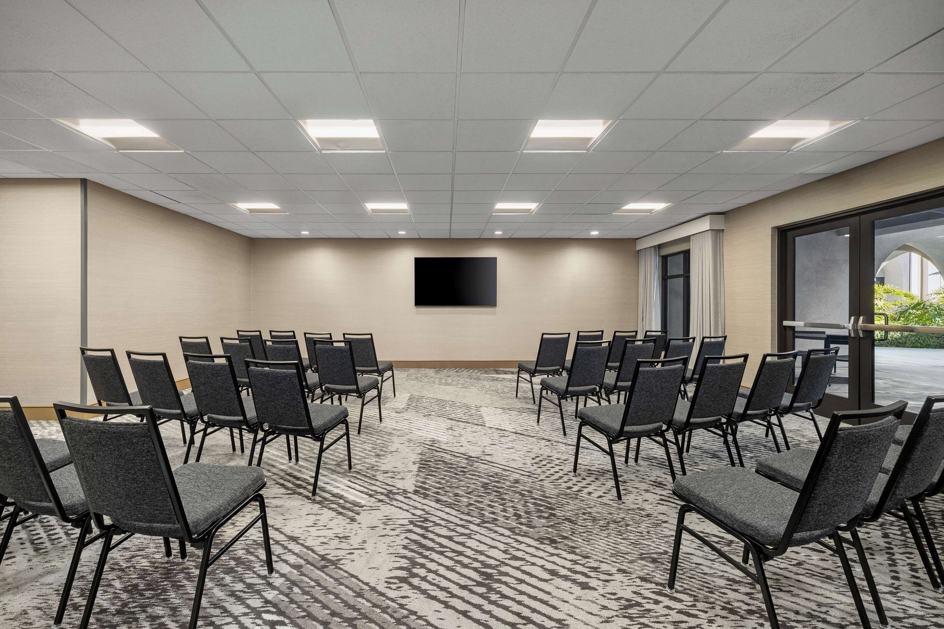Uma sala de conferências com piso carpete, fileiras de cadeiras, uma TV de tela plana na parede e grandes janelas.