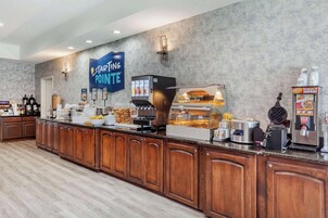 Uma área de café da manhã de hotel com uma placa Starting Pointe, uma máquina de café, uma máquina de waffles e diversas opções de comida à mostra.