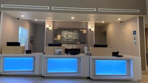 Uma área de recepção de hotel com três balcões, um painel digital grande e uma televisão na parede.