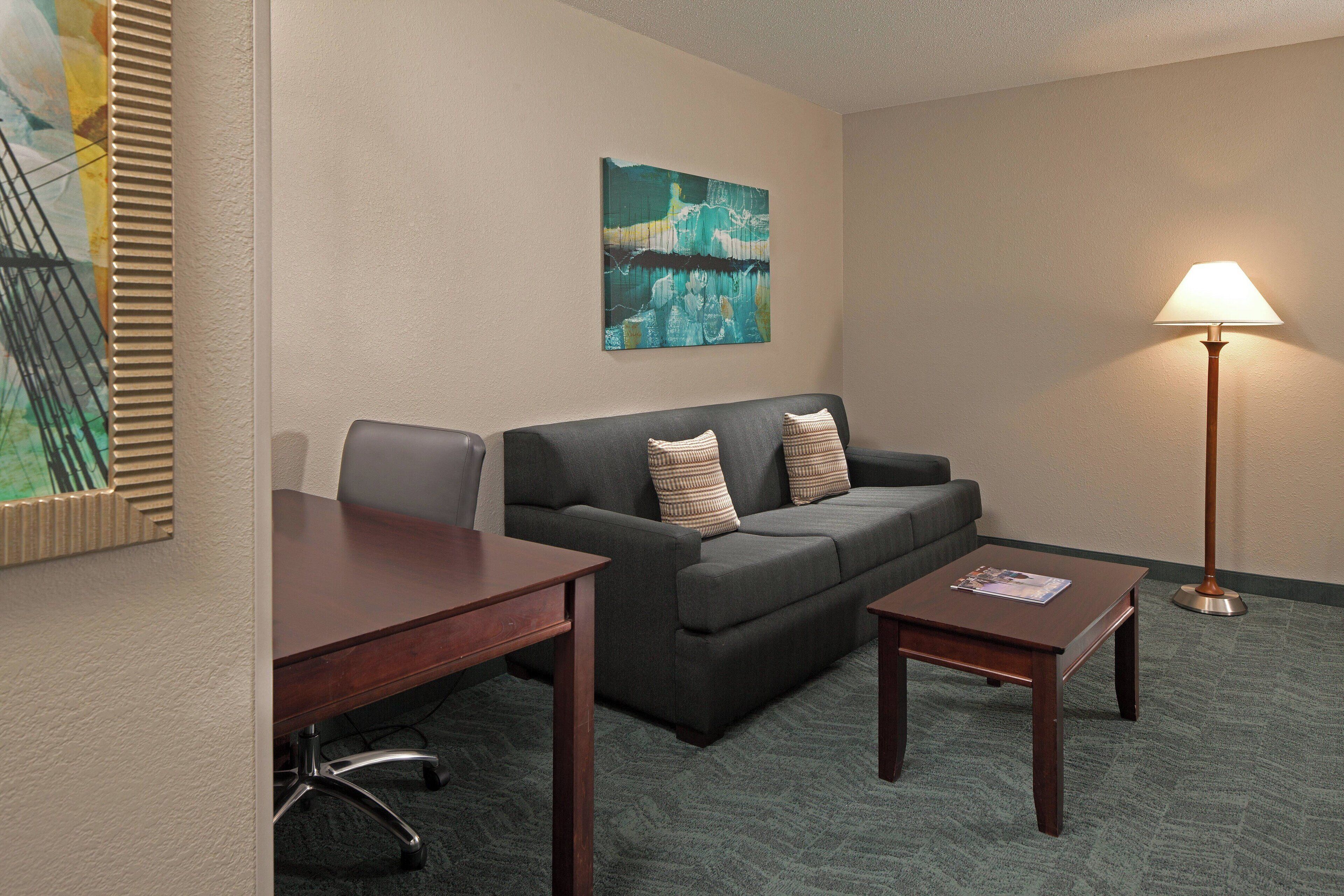 Quarto de hotel com sofá cinzento, mesa de centro de madeira, luminária e um quadro na parede.