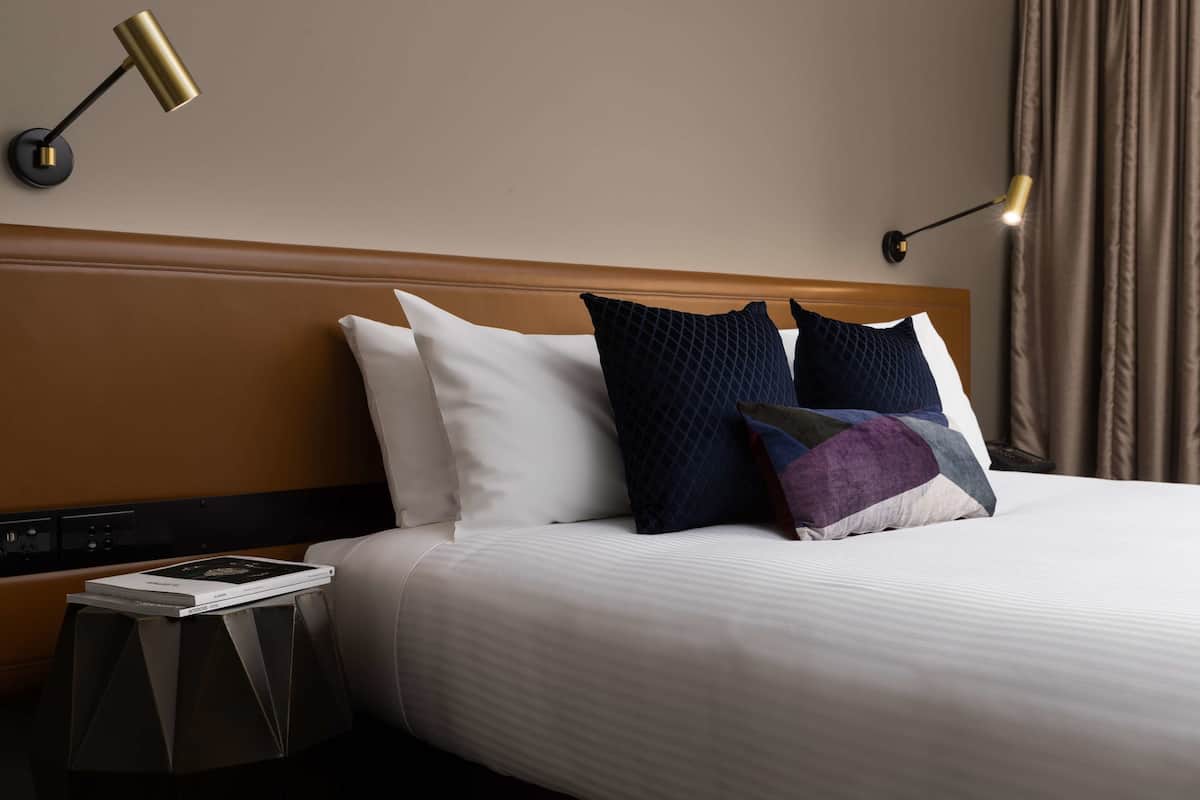 Cama bem arrumada com almofadas brancas e estampadas, mesa de cabeceira com um livro e duas luminárias fixadas na parede.
