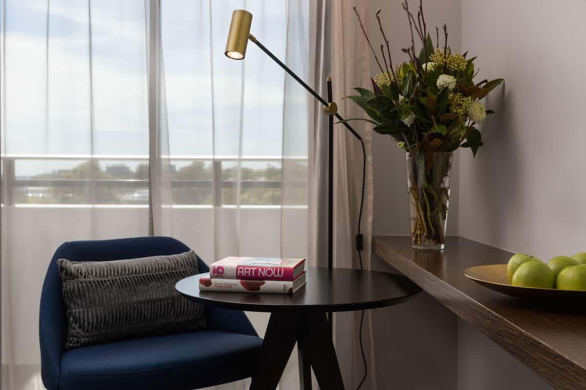 Uma sala de estar moderna com um sofá azul, uma mesinha redonda com um livro, um abajur, um vaso com flores e um prato de maçãs verdes.