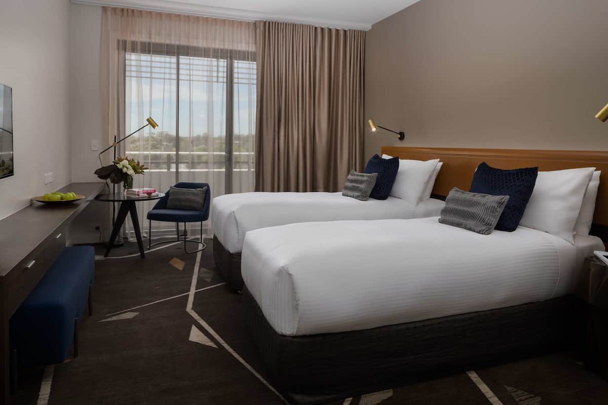 Quarto de hotel com duas camas, uma escrivaninha, uma cadeira, televisão e uma janela grande com cortinas.