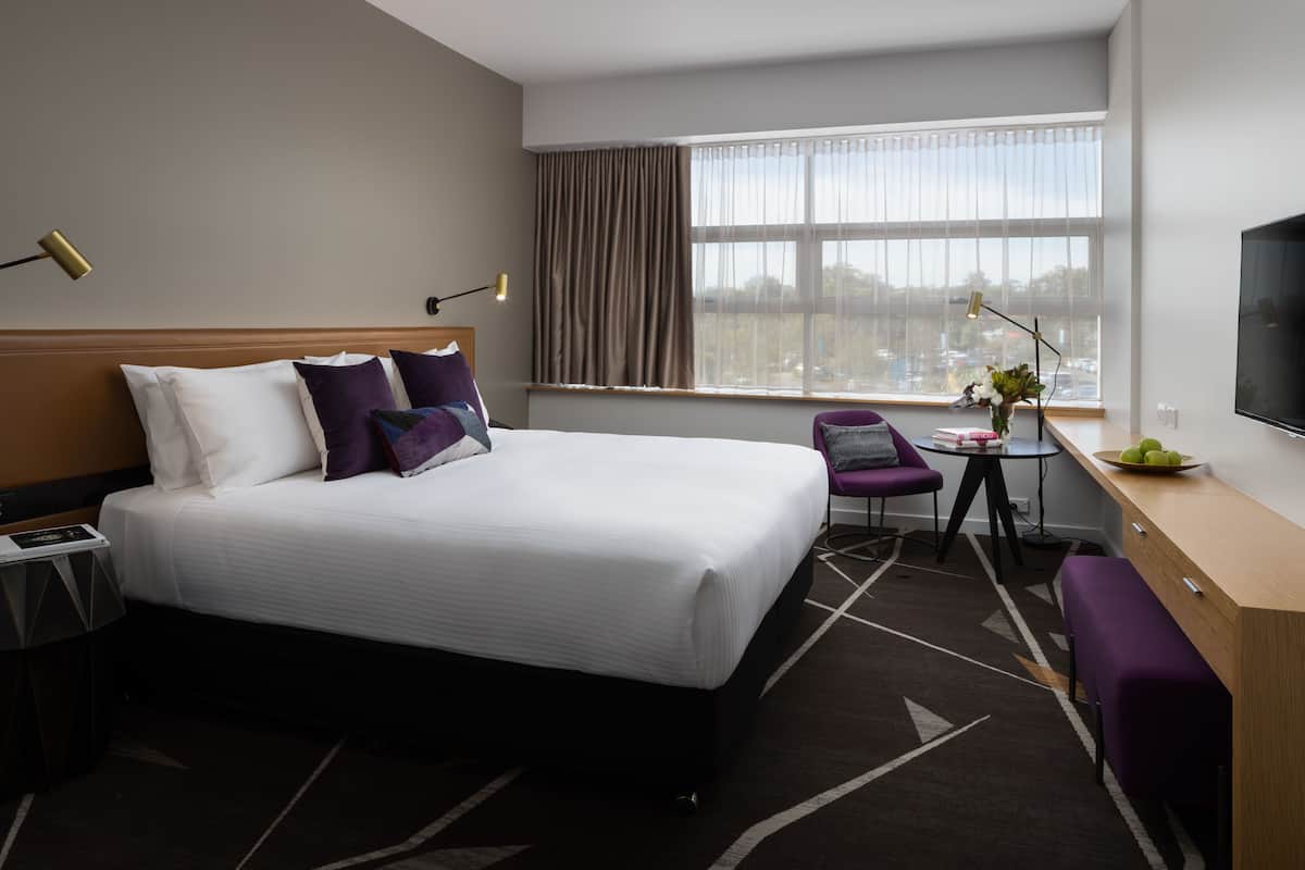 Quarto de hotel com uma cama grande, uma escrivaninha, uma cadeira, uma televisão e uma janela com cortinas.