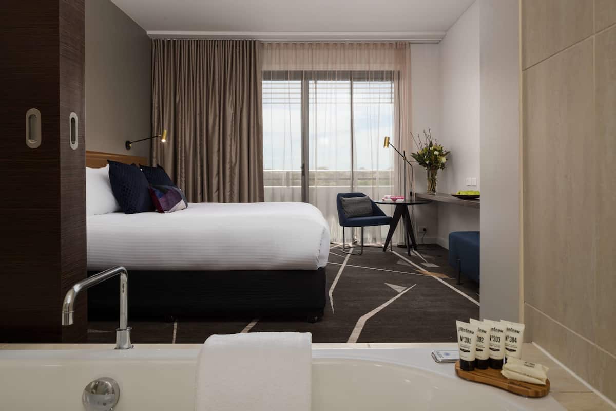 Quarto de hotel com cama, escrivaninha, cadeira, banheira e janela com cortinas.
