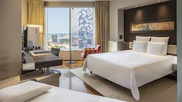 Quarto de hotel com uma cama grande, uma escrivaninha com um computador, uma cadeira e vista para a cidade através de uma janela com padrão geométrico.
