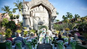 Uma cerimônia de casamento com convidados sentados em cadeiras adornadas com fitas verdes, uma noiva em vestido branco e um noivo em terno escuro.