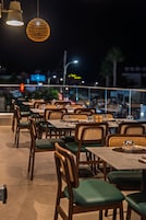 Uma área de refeições ao ar livre com mesas e cadeiras, luminárias de rua e vista da cidade à noite.