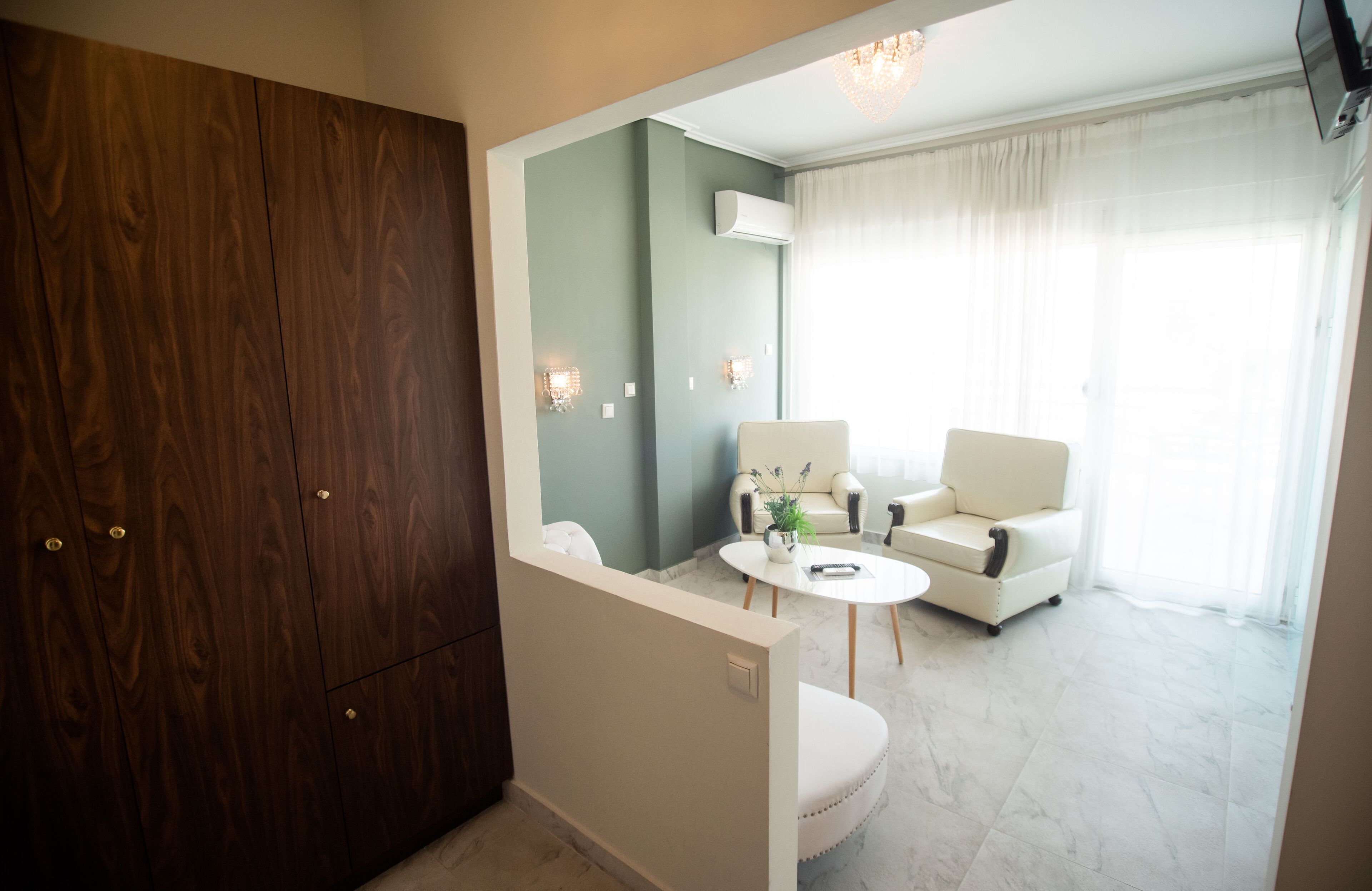 Quarto moderno com guarda-roupa de madeira, uma mesa branca redonda, duas poltronas brancas e uma grande janela com cortinas translúcidas.