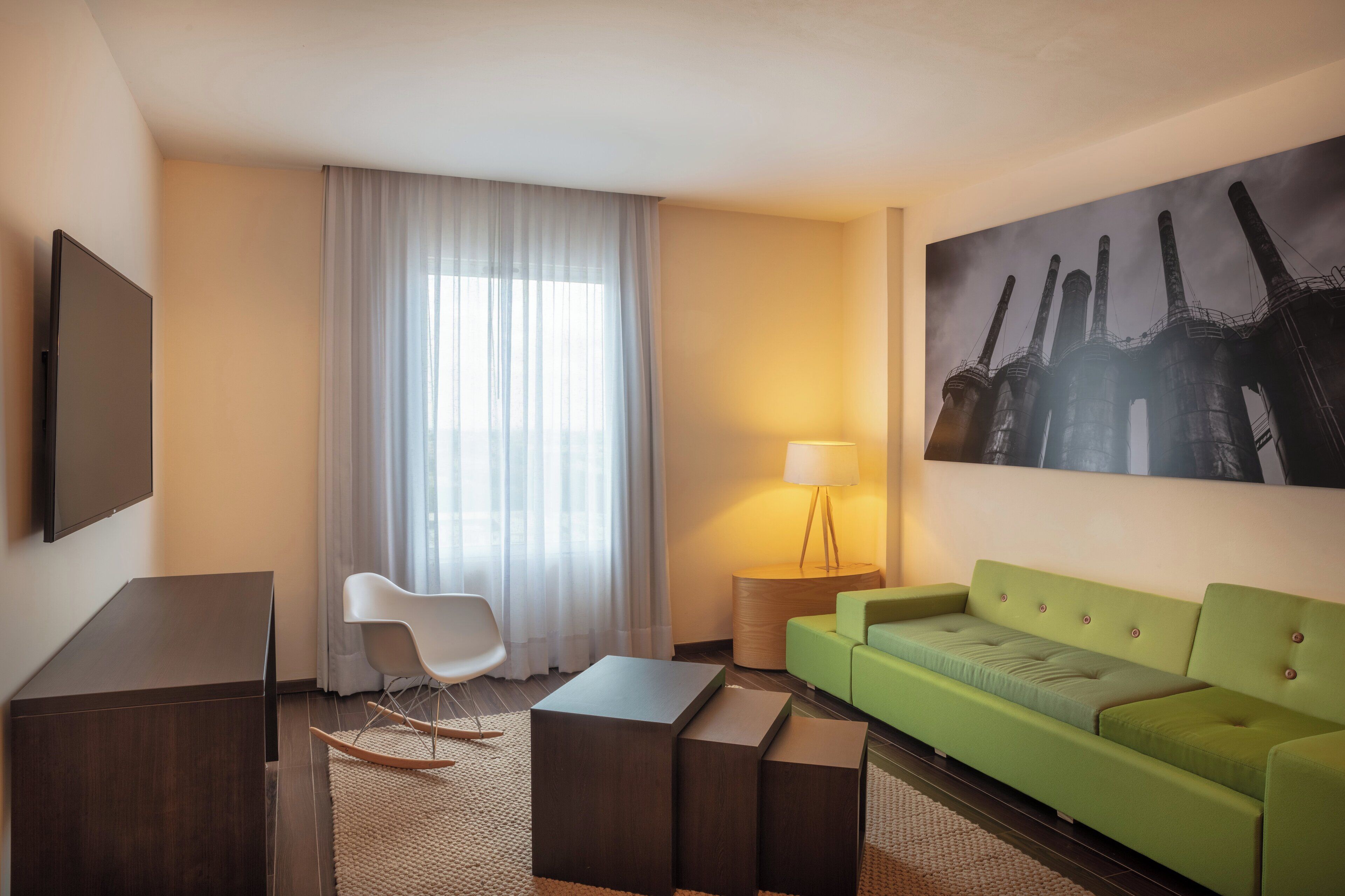 Quarto de hotel com sofá verde, cadeira branca, televisão de tela plana, abajur e pintura de chaminés industriais.