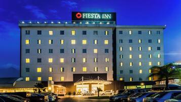 Um hotel de vários andares com uma placa grande e visível com a inscrição "Fiesta Inn" no topo. O edifício possui um amplo estacionamento com vários carros estacionados na frente.