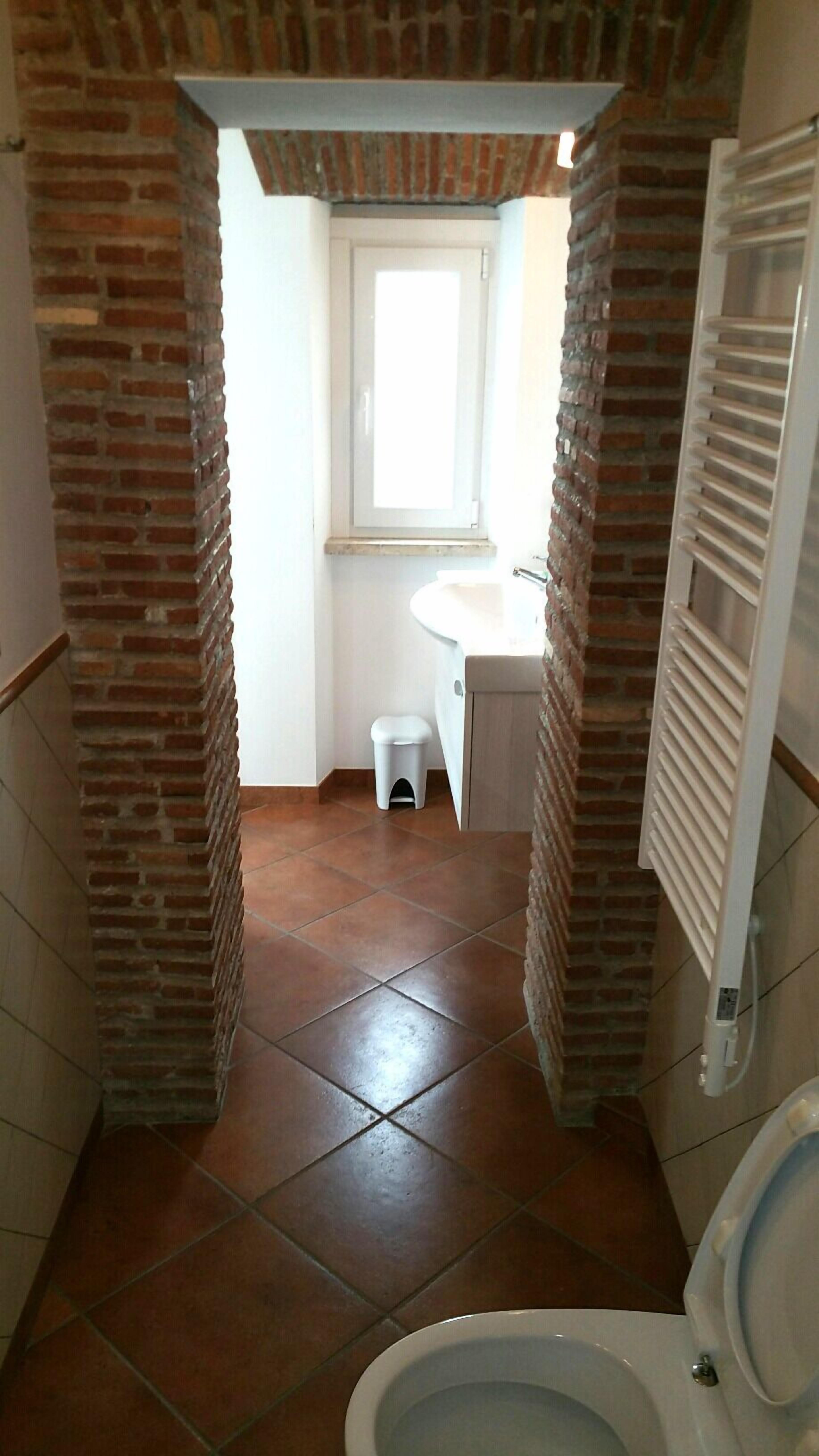 Casa de banho com sanita branca, um banquinho pequeno branco e uma janela.