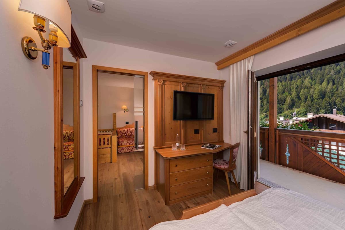 Quarto com penteadeira de madeira, televisão na parede e varanda com vista para uma floresta.
