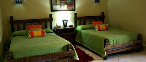Duas camas de madeira com roupa de cama verde e almofadas vermelhas, uma mesa de cabeceira com abajur e um quadro abstrato emoldurado na parede.