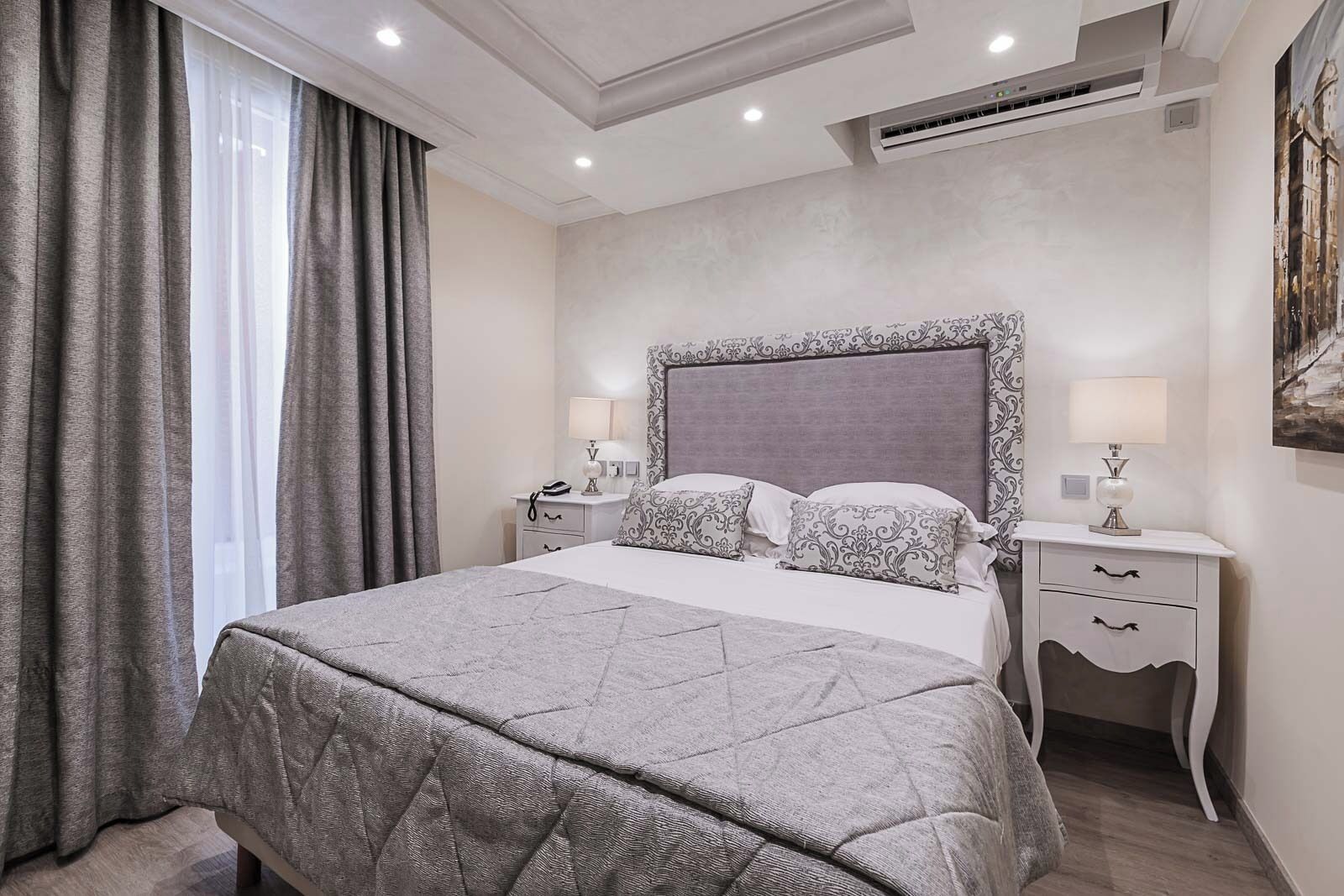 Quarto moderno com uma cama grande, mesas de cabeceira, um criado-mudo, um quadro na parede e um ar condicionado.