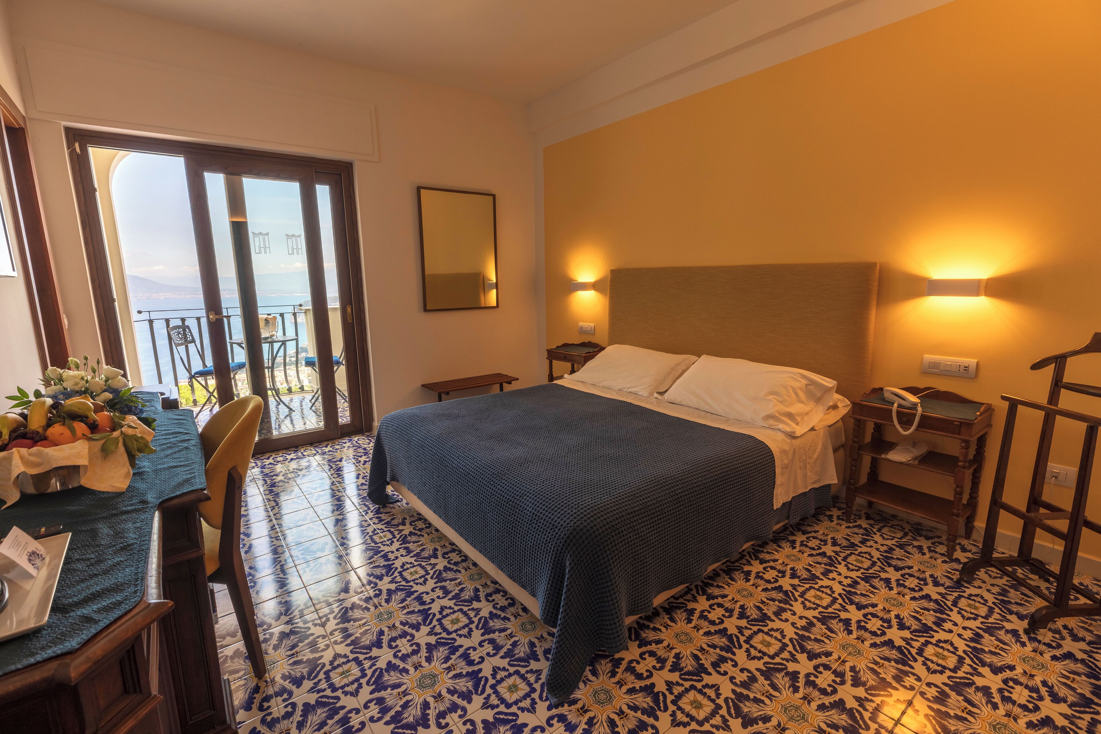 Quarto de hotel com uma cama, uma escrivaninha com cadeira, uma mesa com frutas e flores, uma varanda com vista e ladrilhos estampados no chão.
