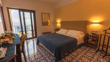 Quarto de hotel com uma cama, uma escrivaninha com cadeira, uma mesa com frutas e flores, uma varanda com vista e ladrilhos estampados no chão.